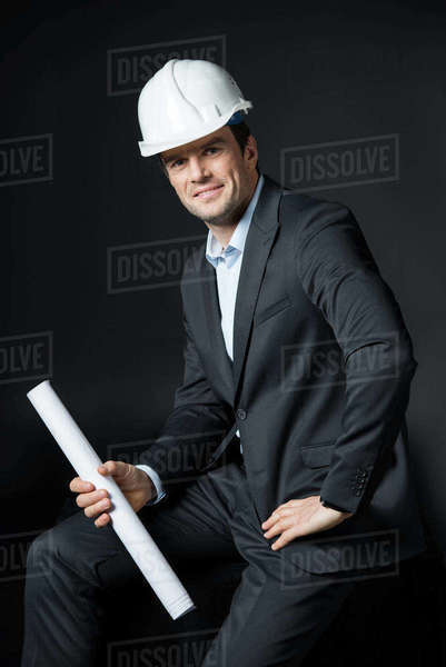 Smiling handsome male engineer in helmet holding blueprint and looking ...