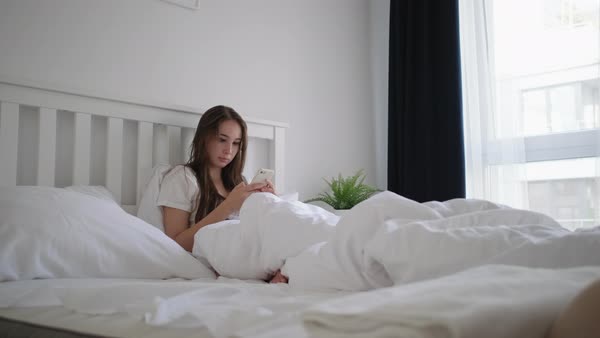 Female Is Lying In Bed In Morning And Looking At Screen Of Her Cell