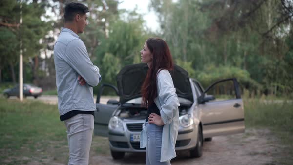 Unsure young man and woman talking standing in forest with broken car. Side view portrait of ...