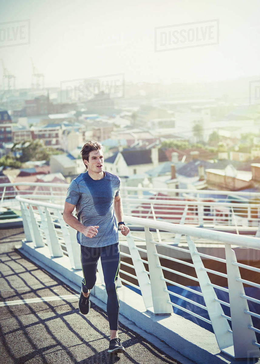 Sweaty male runner running on sunny urban footbridge at sunrise ...