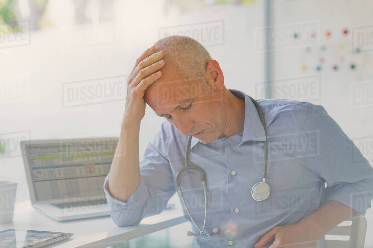 Tired, stressed male doctor with head in hands in doctor’s office ...