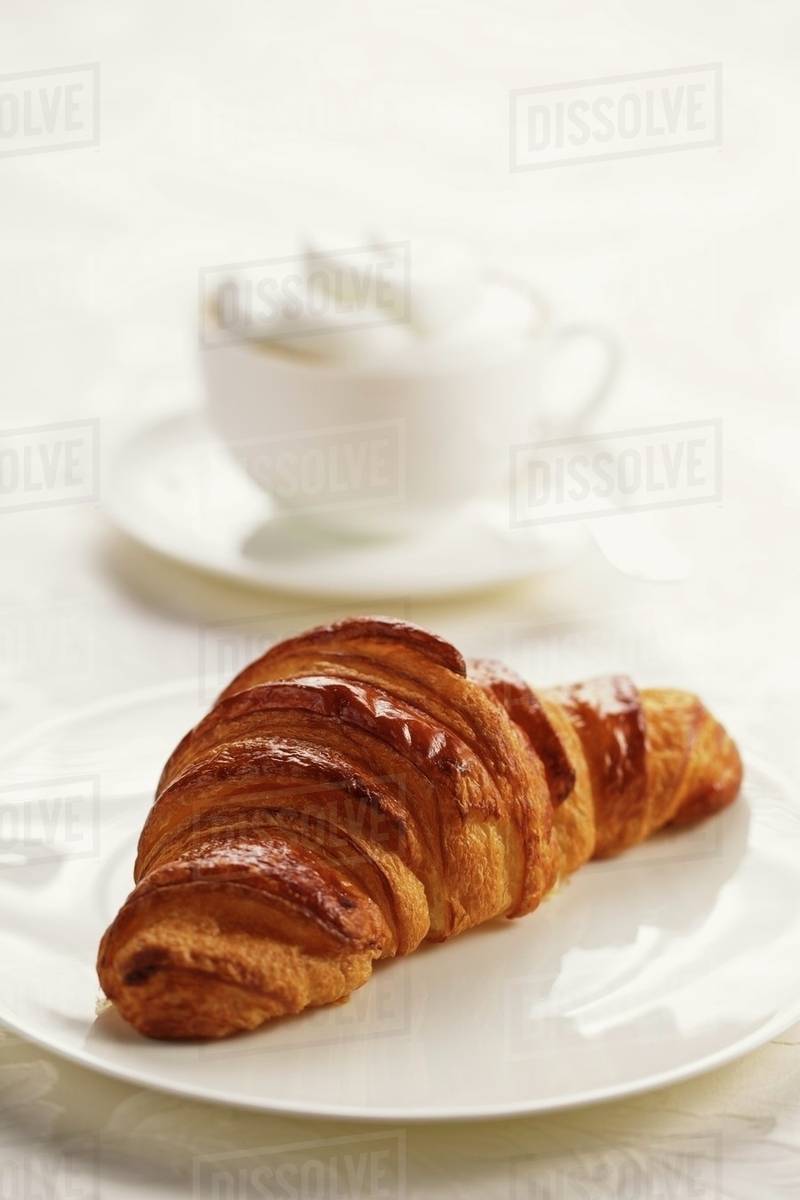A croissant on a plate with a cappuccino in the background - Stock ...