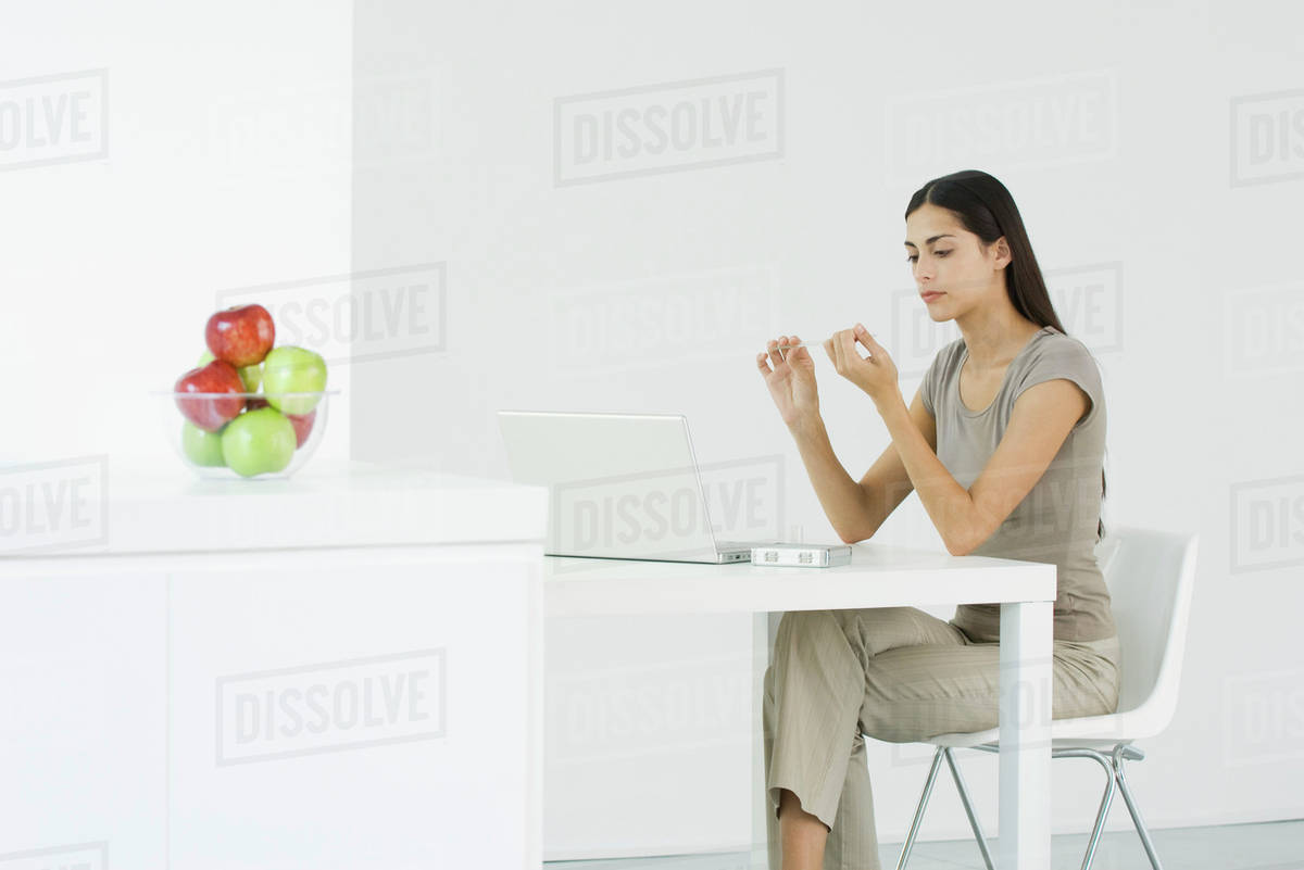 Woman sitting at table in front of laptop computer, filing nails ...