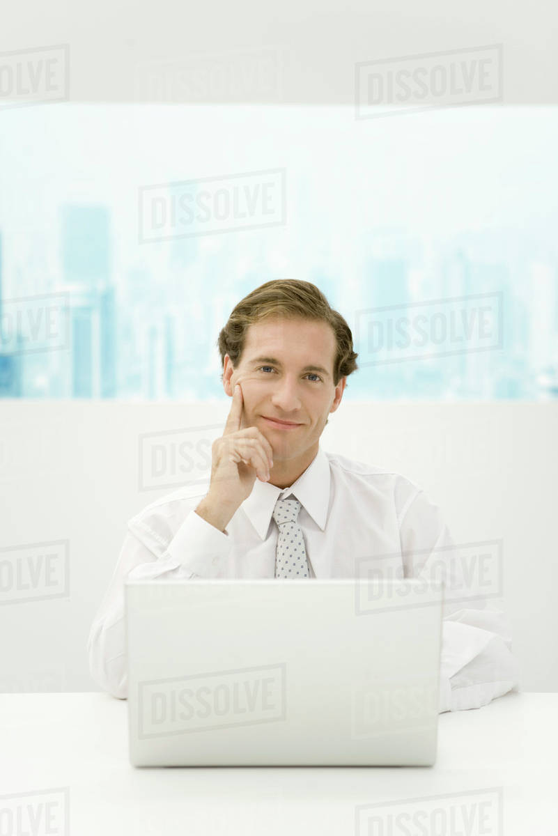Businessman sitting with laptop computer, holding head, smiling at ...
