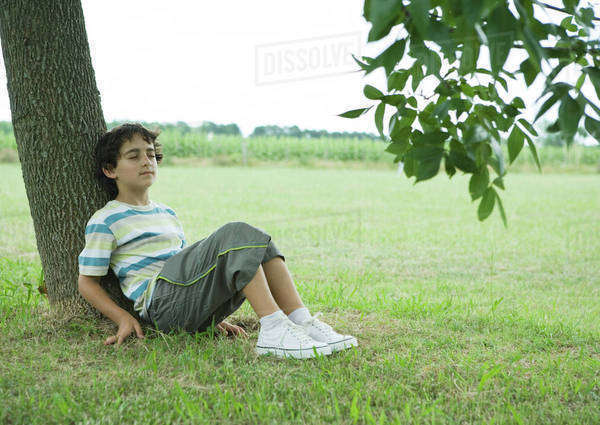 Boy sitting on ground, leaning against tree trunk - Royalty-free Stock ...