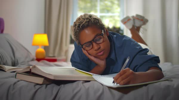 African-American teen school girl doing homework lying on bed at home ...