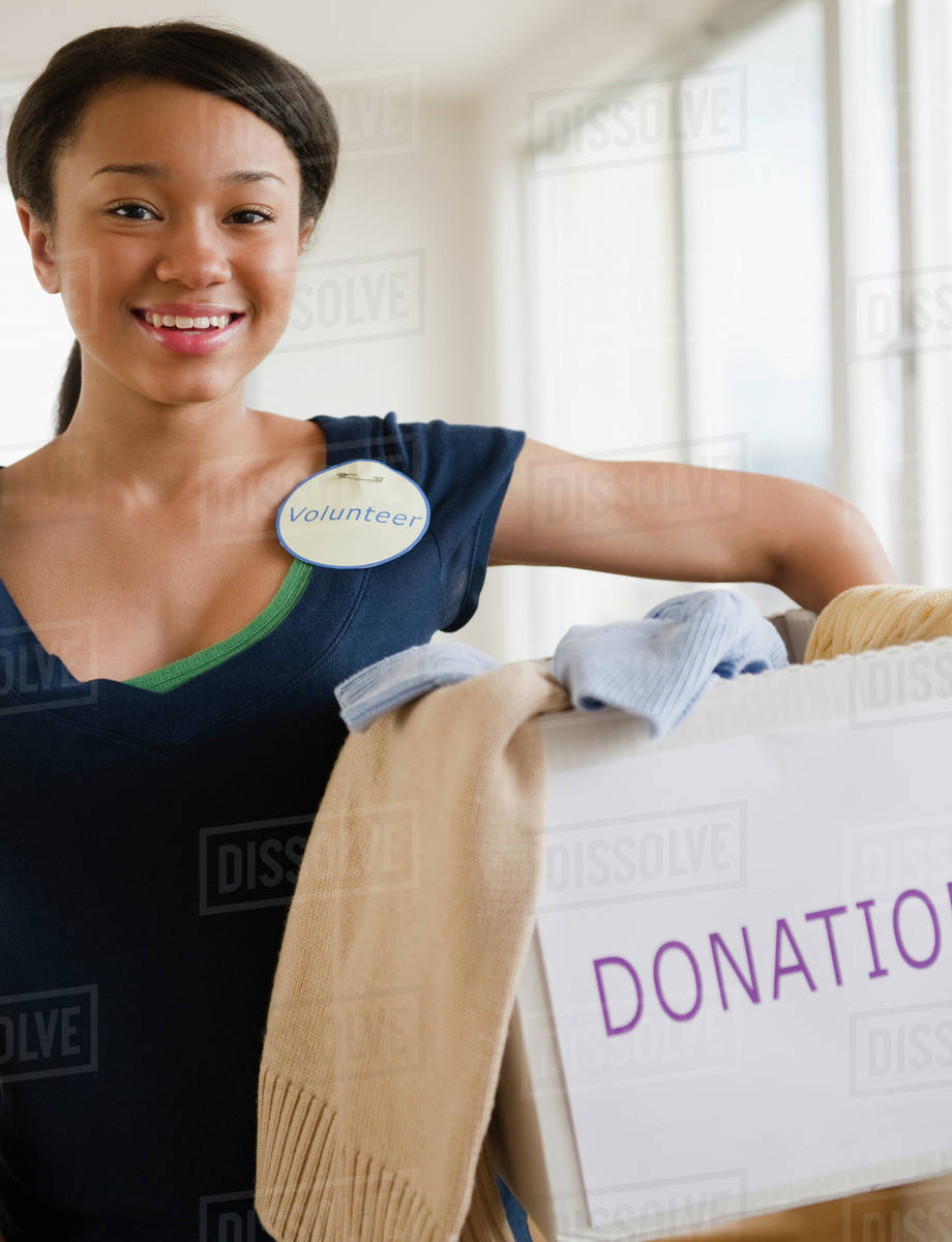 Smiling mixed race teenage girl wearing 'Volunteer' pin and holding box ...