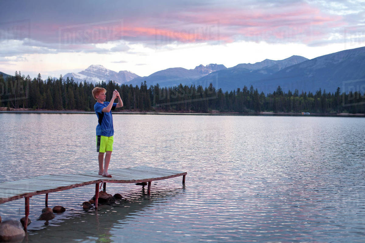 Boy standing on wooden dock photographing lake - Royalty-free Stock ...