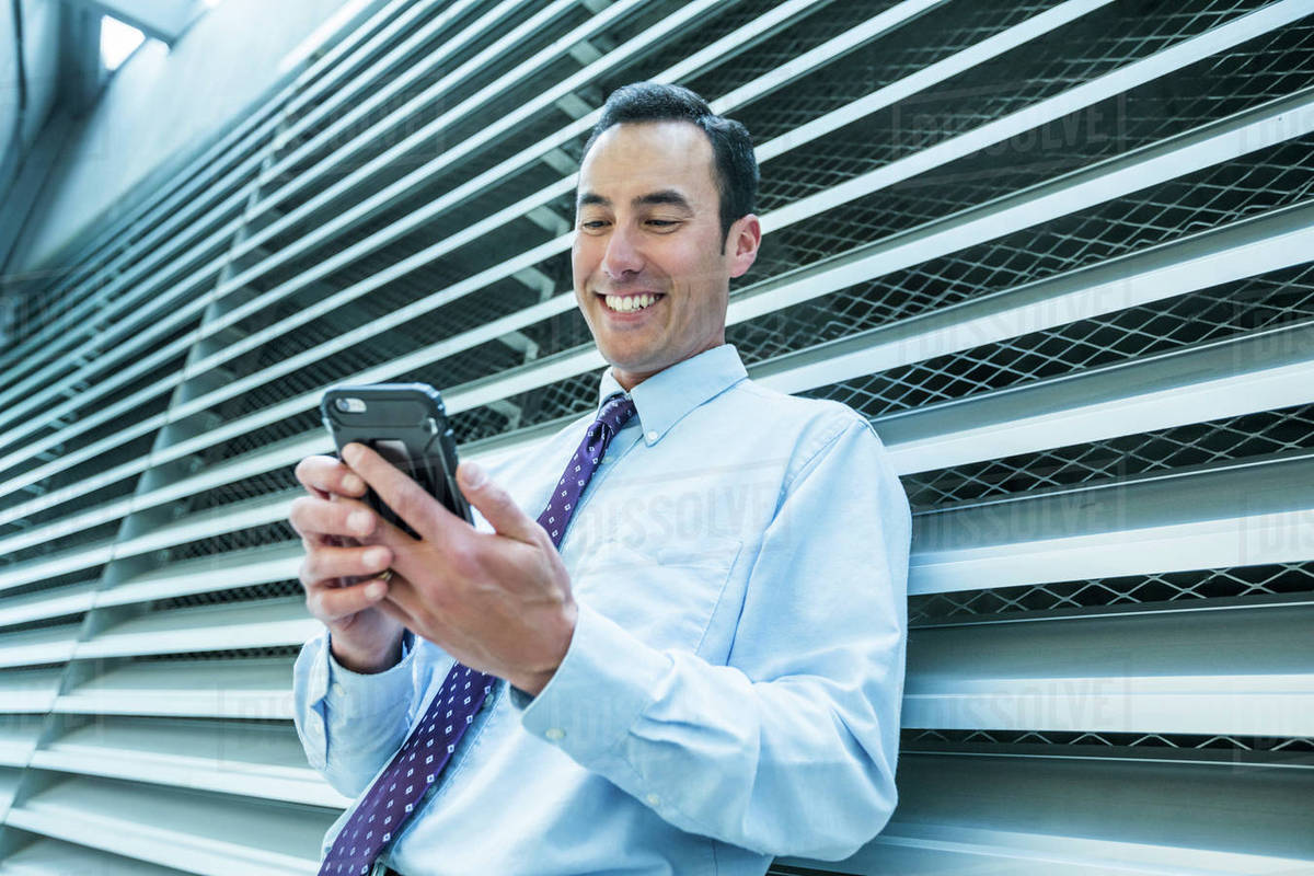 Smiling Mixed Race businessman leaning on wall texting on cell phone ...