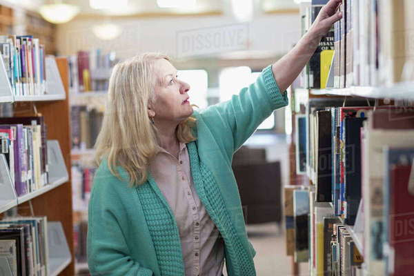 Curious Caucasian woman reaching for book in library - Stock Photo ...