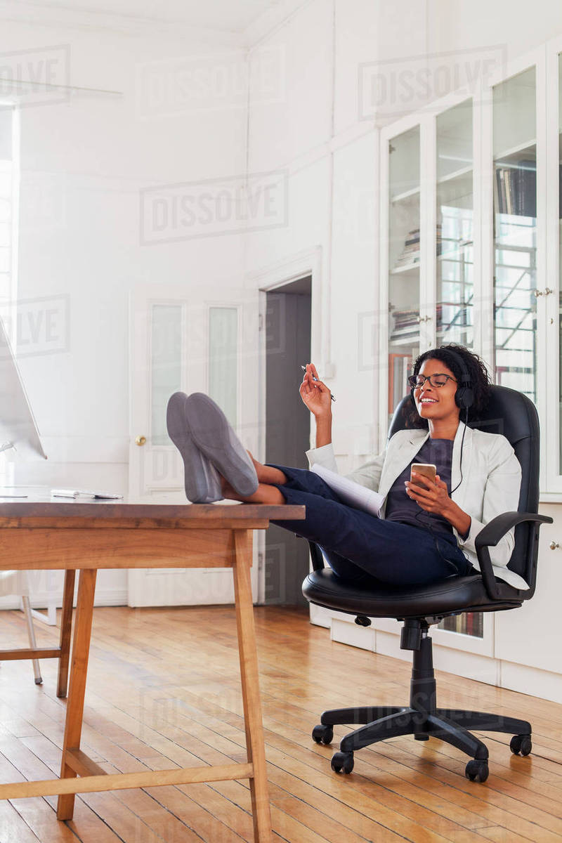 Mixed Race woman with feet up listening to cell phone with headphones ...