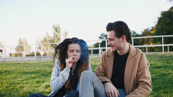 Happy young hipster couple sitting on grass and smoking marijuana joint ...
