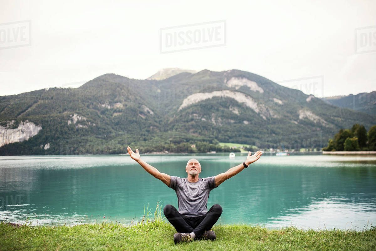 A senior man pensioner sitting by lake in nature, doing yoga exercise ...