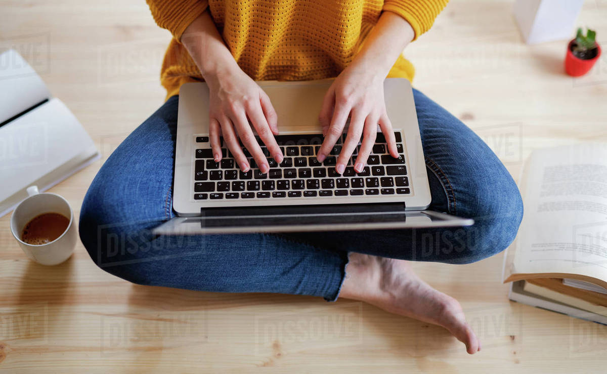 A midsection of young female student sitting on floor using laptop when ...