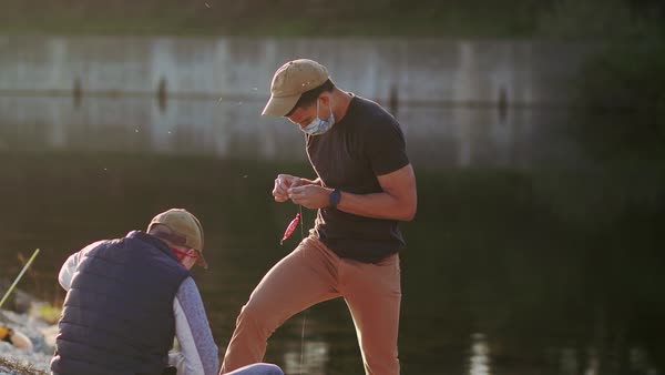 Medium shot of men tying artificial fly to fishing rod string by lake ...