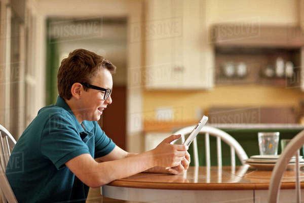 Teenage boy sitting at a kitchen table and looking at a digital tablet ...