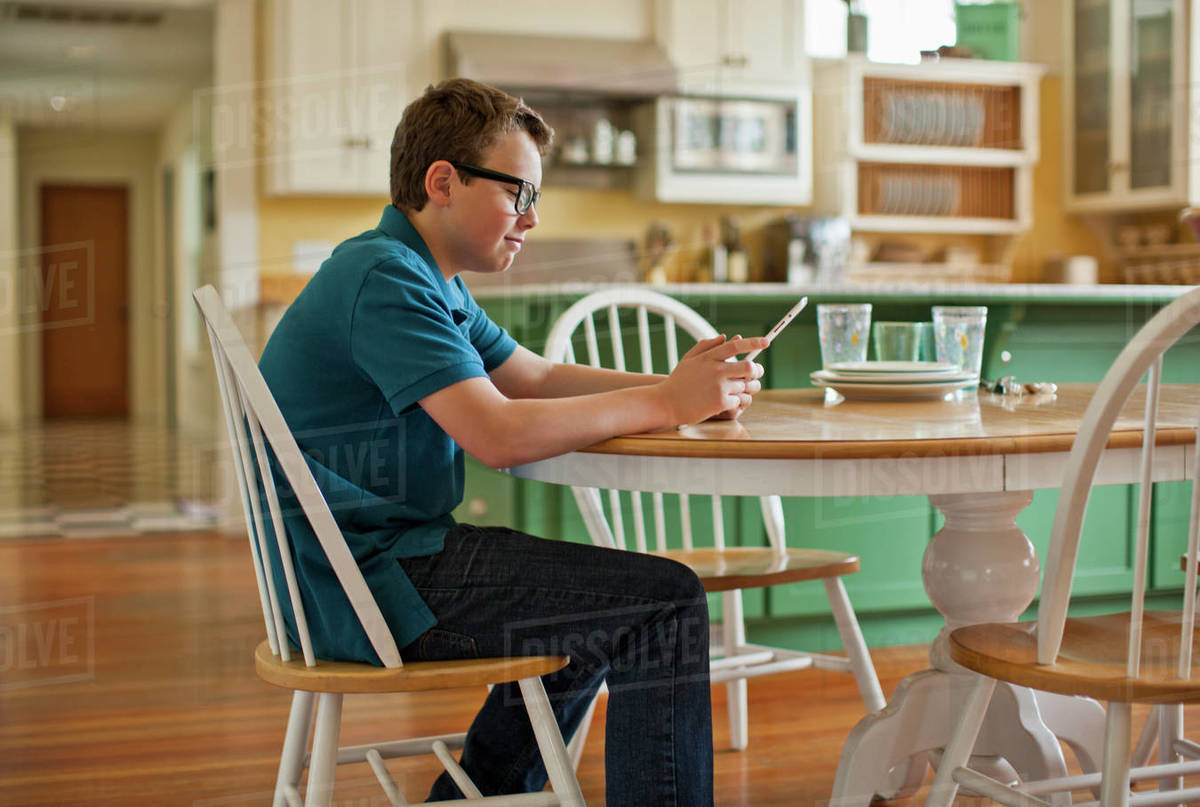 Teenage boy sitting at a kitchen table and looking at a digital tablet ...