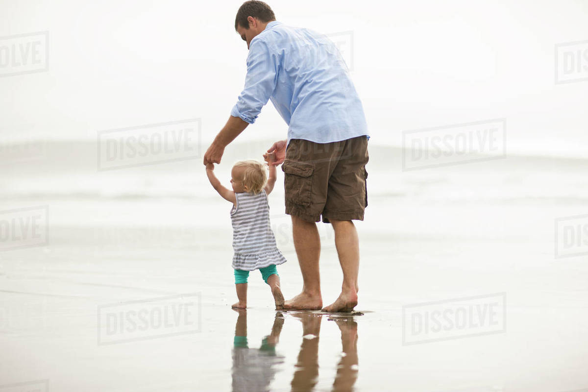 Father helping his baby daughter take her first steps on a beach ...