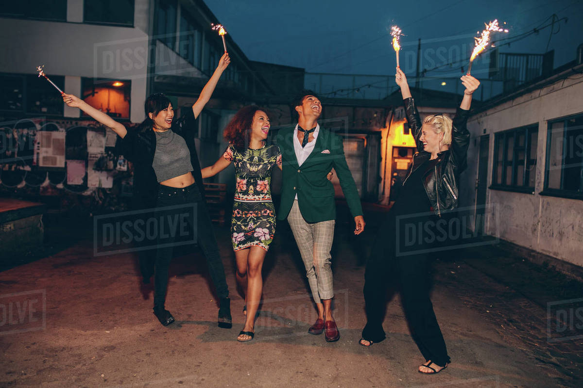 Excited young friends walking on city street at night with fireworks ...