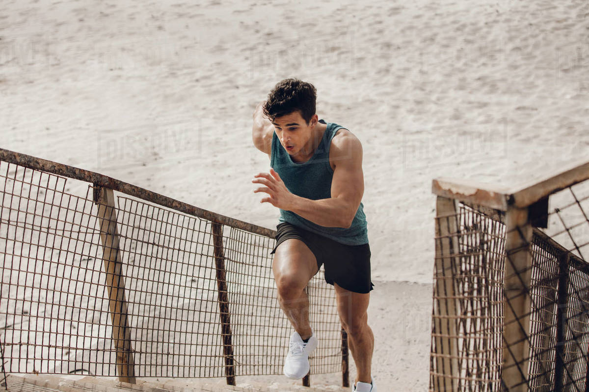 Fit young man running up the steps at beach. Male runner working out on ...