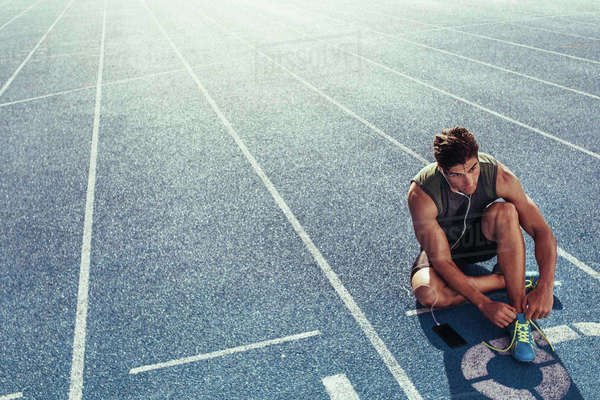 Athlete sitting on a running track tying shoe lace while listening to ...