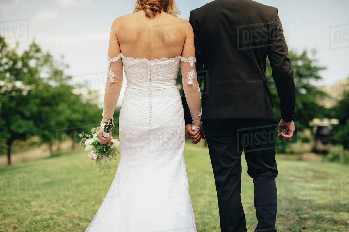 Rear view of bride and groom walking and holding hands on a meadow ...