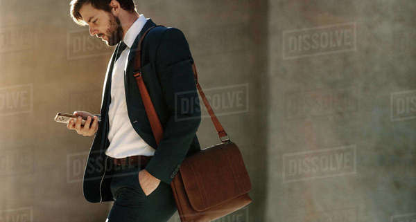 Businessman looking at his mobile phone while walking on street to ...