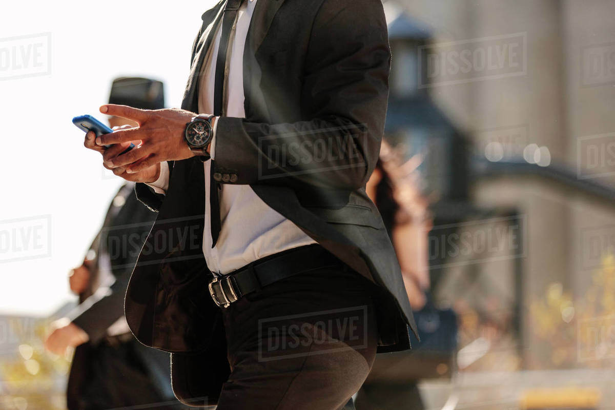 Mid section of a businessman using mobile phone while walking on city ...
