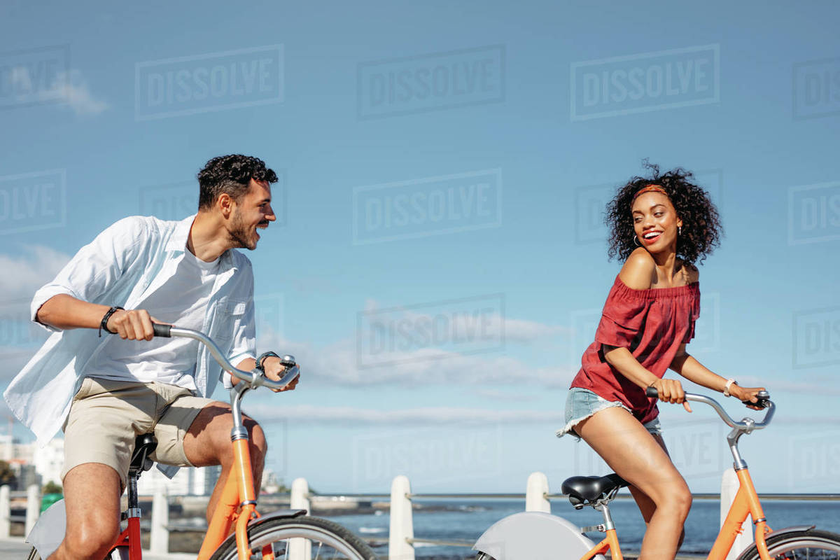 Excited couple riding bicycles in the street with sea in the background ...