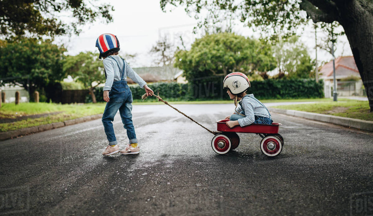Little girl wearing helmet pulling her sister sitting in a wagon cart ...