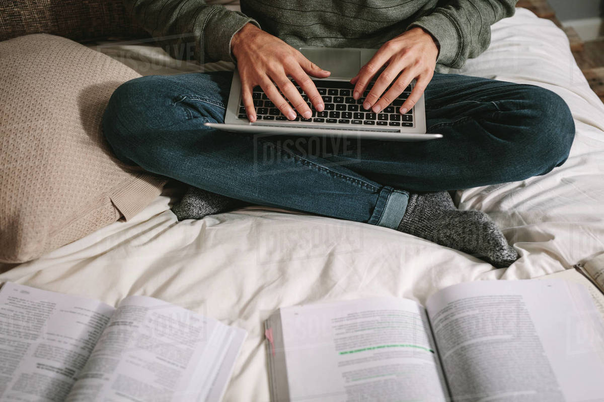 Student using laptop for reference while studying sitting on bed. Young ...