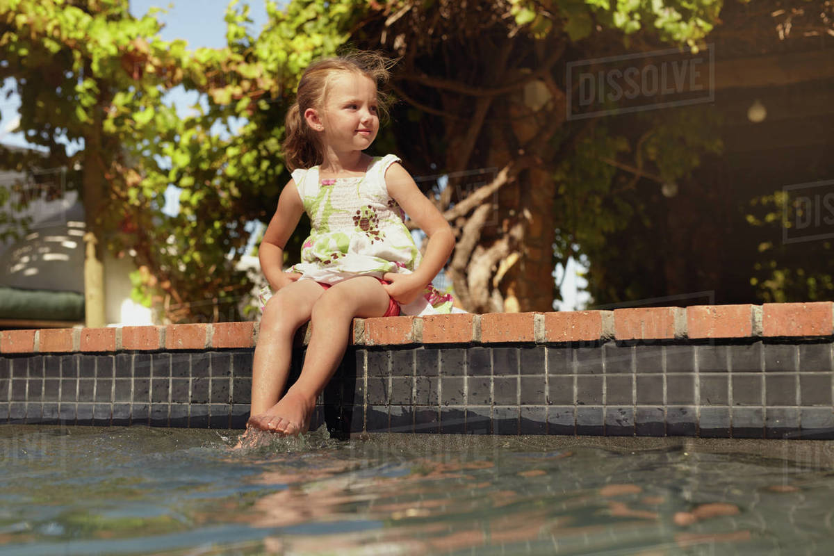 Outdoor shot of a little girl dipping her feet in the pool looking away ...