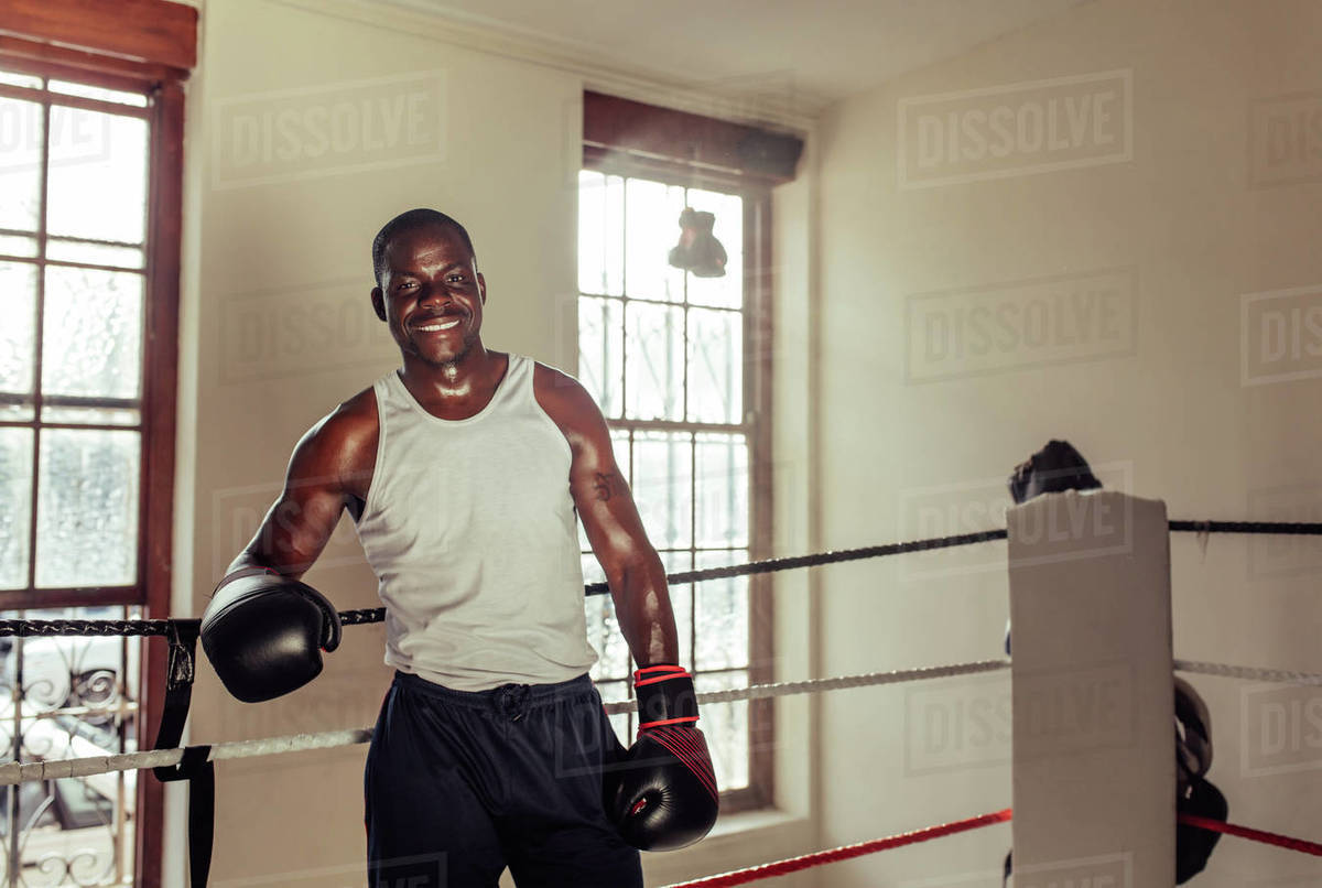 Happy sweaty fit young African boxer leaning back against the ropes in ...