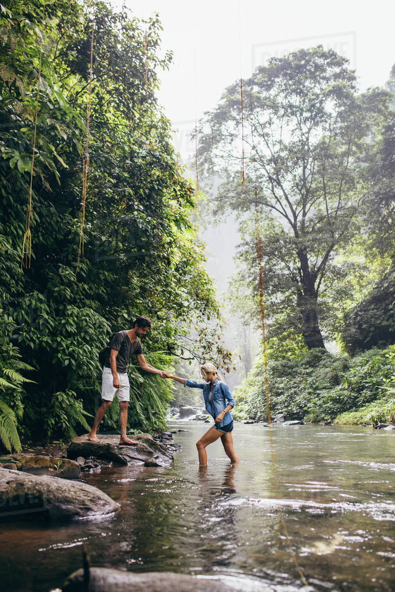 Outdoor shot of young man helping woman crossing stream. Couple in ...