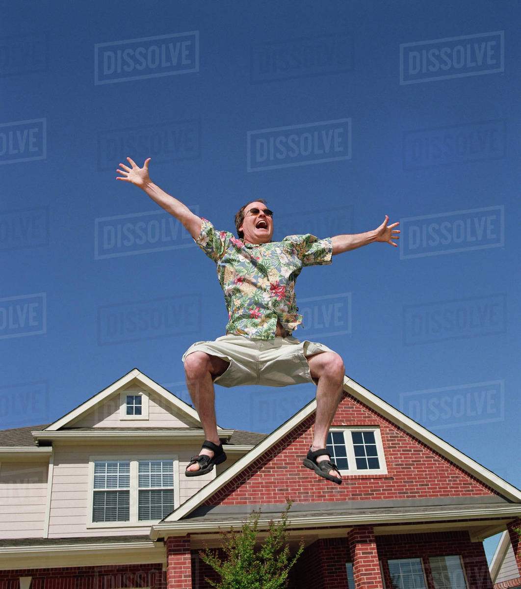 Middle aged man jumping in front of home, striking a pose mid air ...