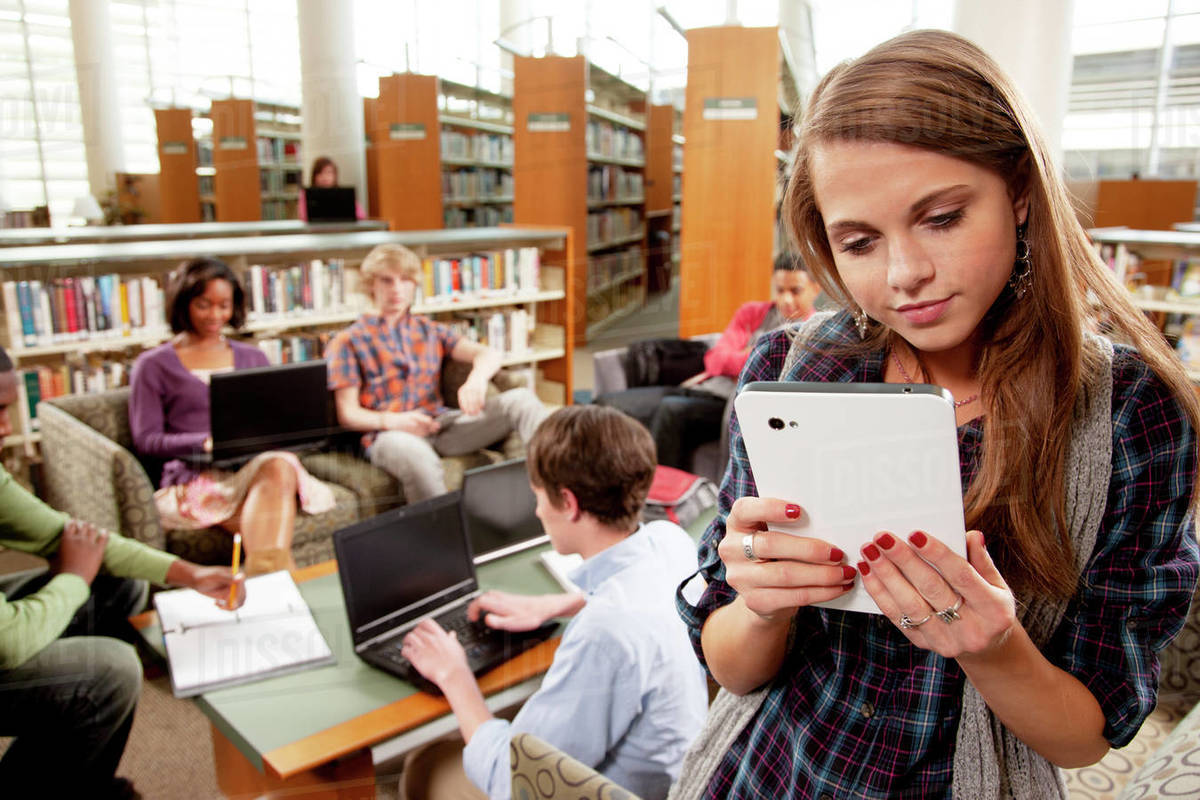 Focused college girl reading on tablet in library with friends working ...