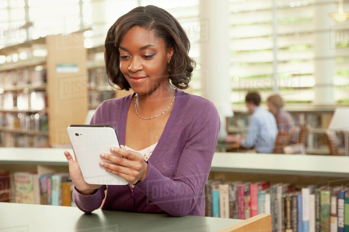 Female student using tablet, leaning against bookshelf in library ...
