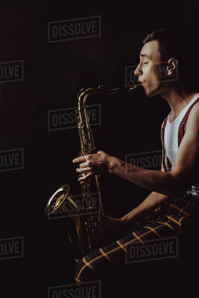 Side view of stylish young saxophonist sitting and playing sax on black ...