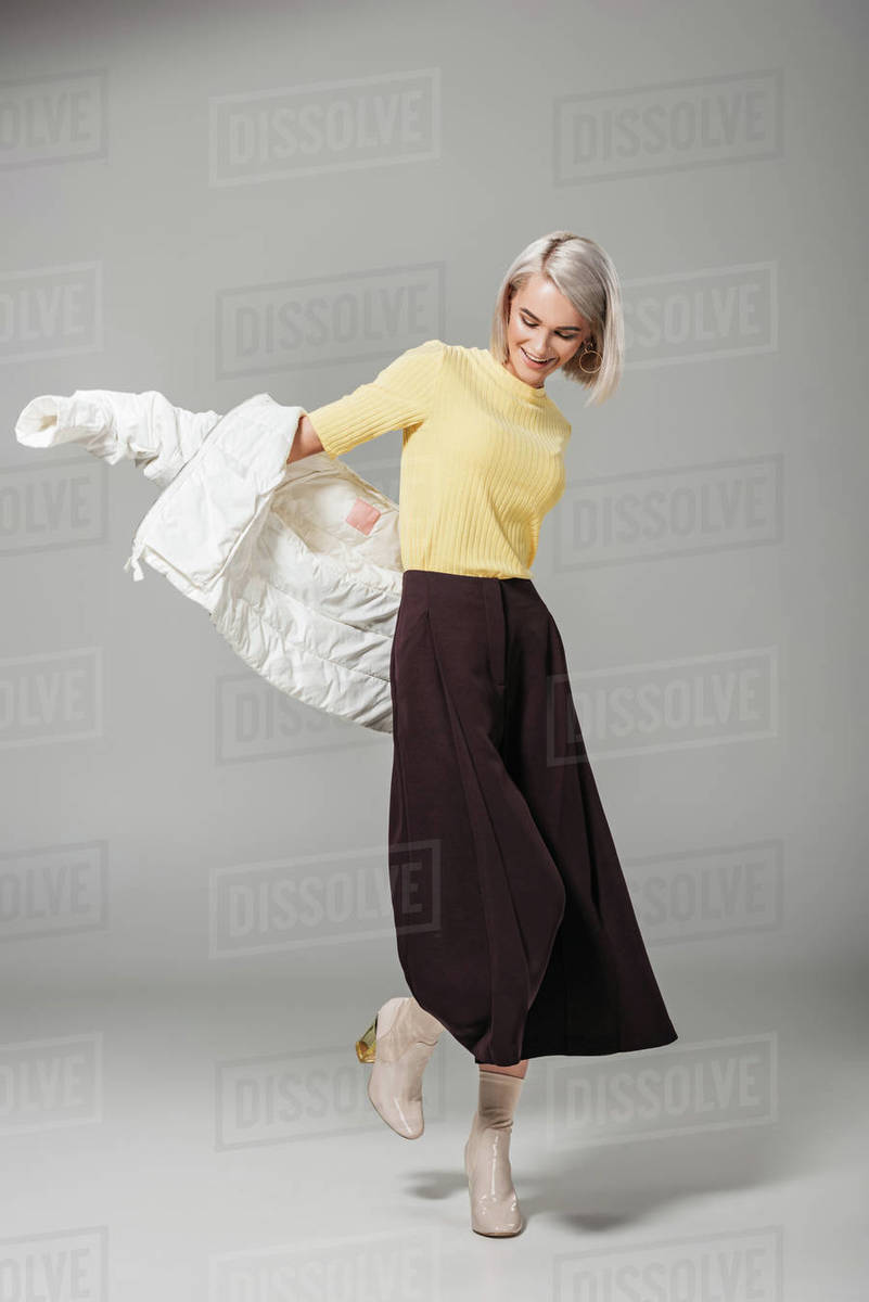 smiling young woman putting on stylish white jacket on grey background ...