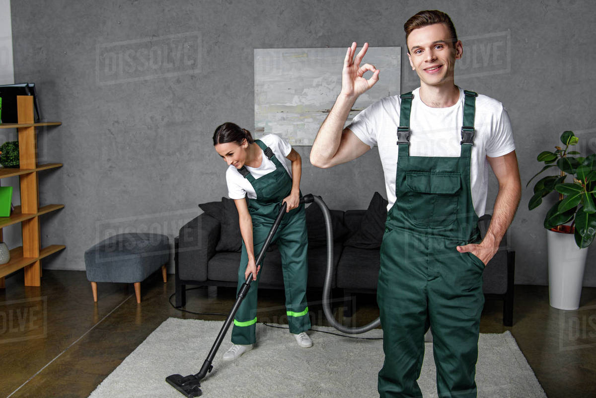 Smiling young cleaning company workers using vacuum cleaner and showing ...