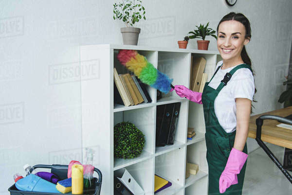 Beautiful young cleaner holding duster and smiling at camera while ...
