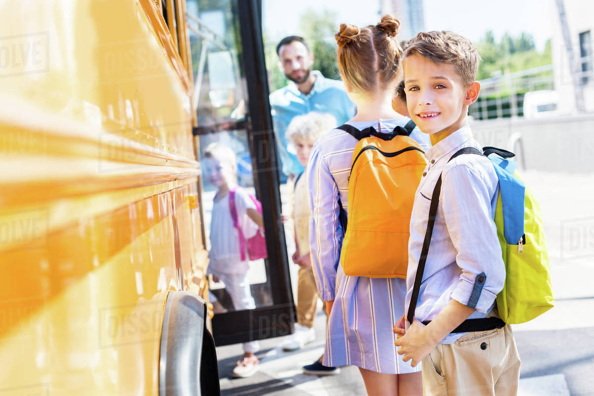 adorable schoolboy entering school bus with classmates while teacher ...