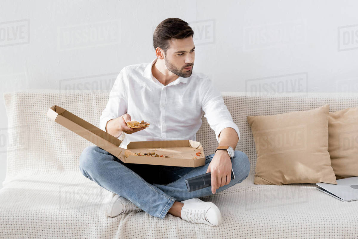 Young man with remote control in hand eating pizza alone at home ...