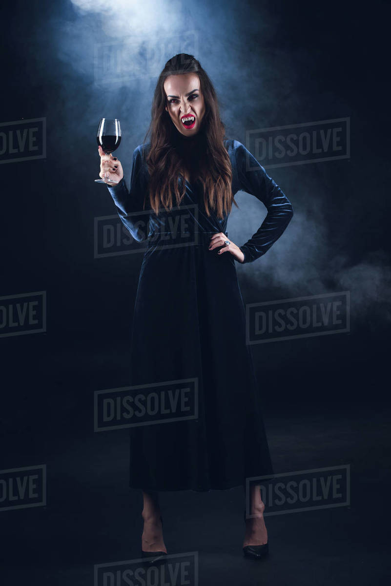 Grim vampire holding wineglass with blood on dark background with smoke ...