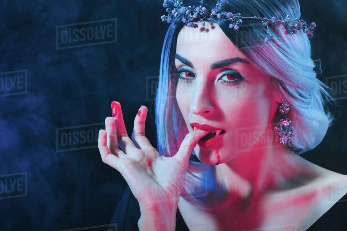 Attractive vampire licking bloody fingers on dark background with smoke ...