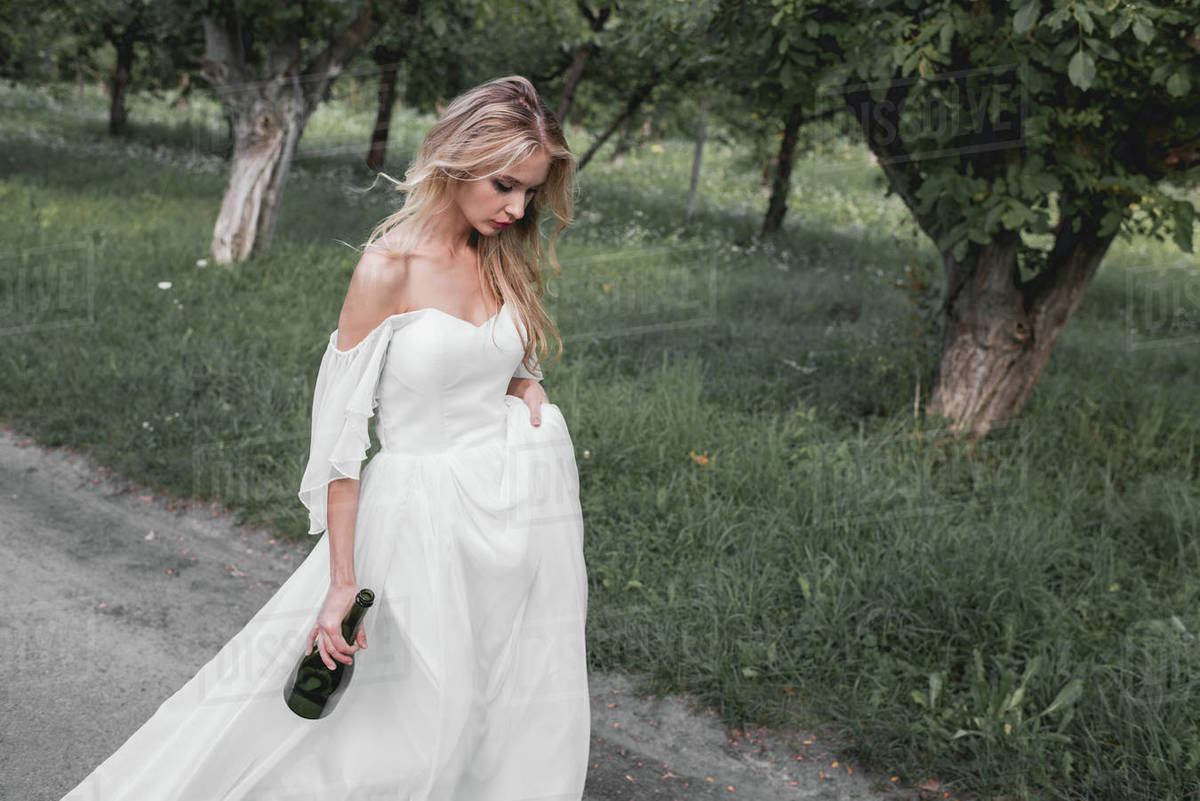 upset young bride in wedding dress holding bottle of wine and walking ...