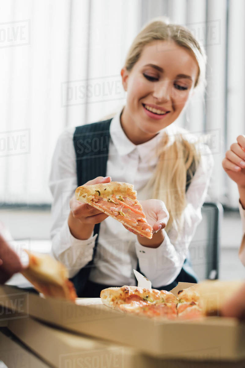 Smiling young businesswoman eating pizza with colleagues in office ...