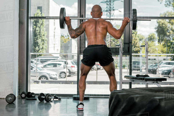 rear view of athletic young shirtless man lifting barbell in gym ...