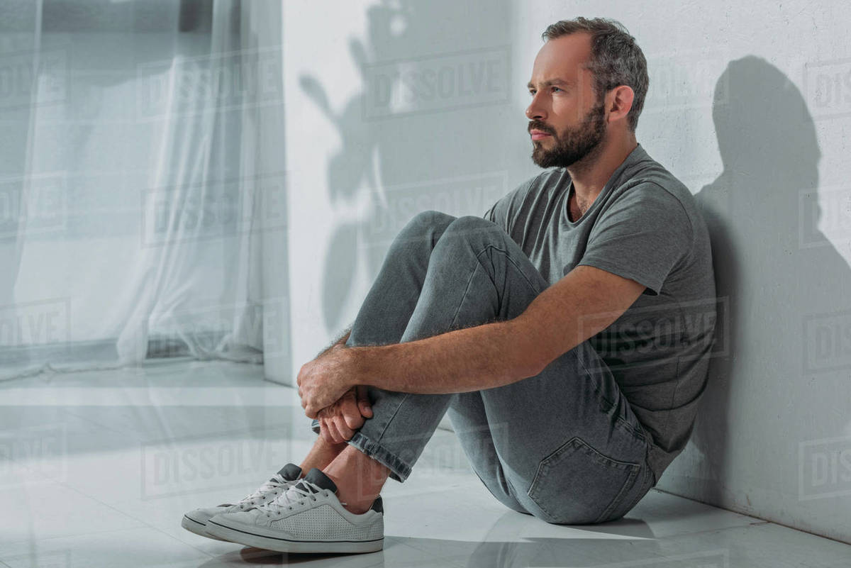 Side view of sad bearded mid adult man sitting on floor and looking ...