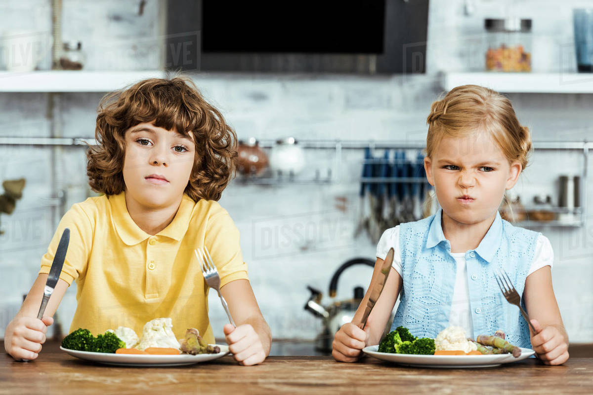 Cute unhappy kids holding cutlery and looking at camera while sitting ...